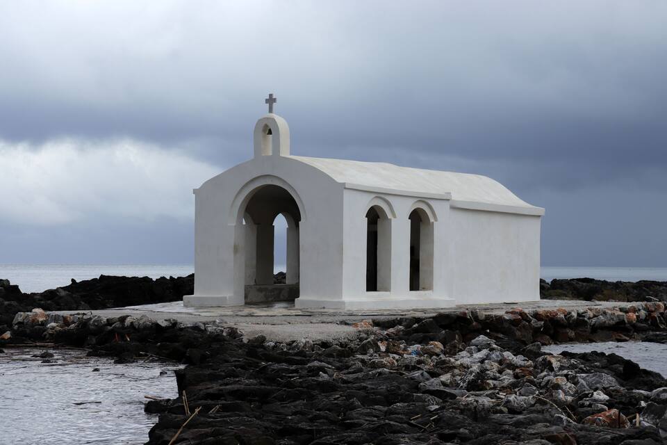 Uma igreja branca com uma cruz no topo, situada numa costa rochosa com o oceano ao fundo.