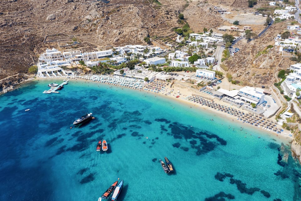 Uma vila costeira com edifícios brancos, uma praia e barcos em águas azul-claras.