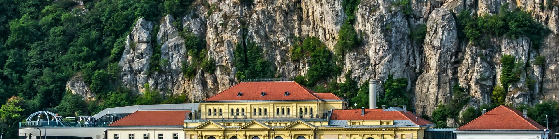 Um edifício histórico com fachada amarela e telhado vermelho, localizado próximo a uma linha de trem, com montanhas ao fundo.