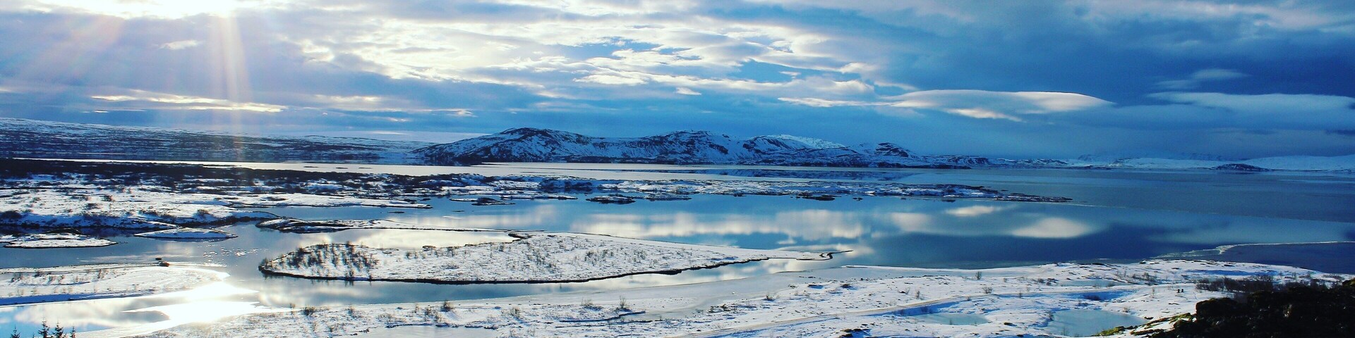 Paisagem nevada com um lago, montanhas e céu parcialmente nublado.