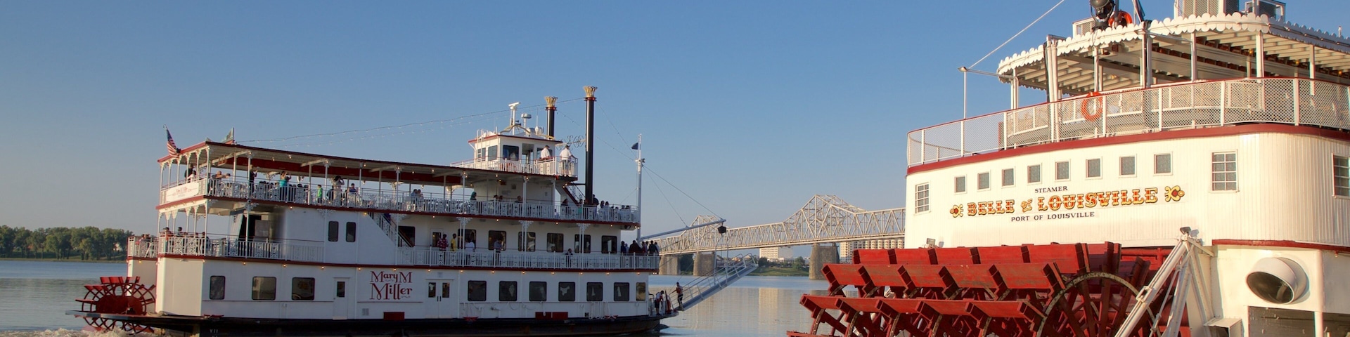 Um barco a vapor atracado em um cais, com uma grande roda de pás e uma ponte ao fundo.