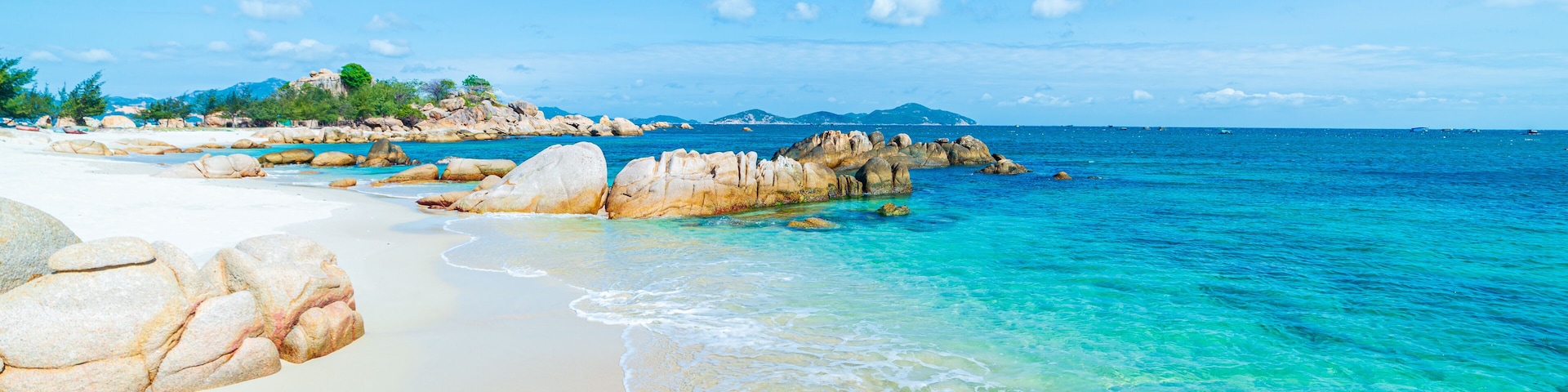 Uma praia com pedras grandes, água turquesa transparente e montanhas ao longe.