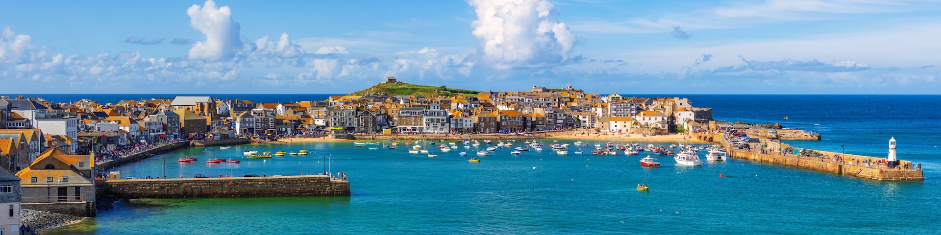 Uma cidade costeira com edifícios coloridos, um porto cheio de barcos e uma colina imponente com uma estrutura semelhante a um castelo ao fundo.