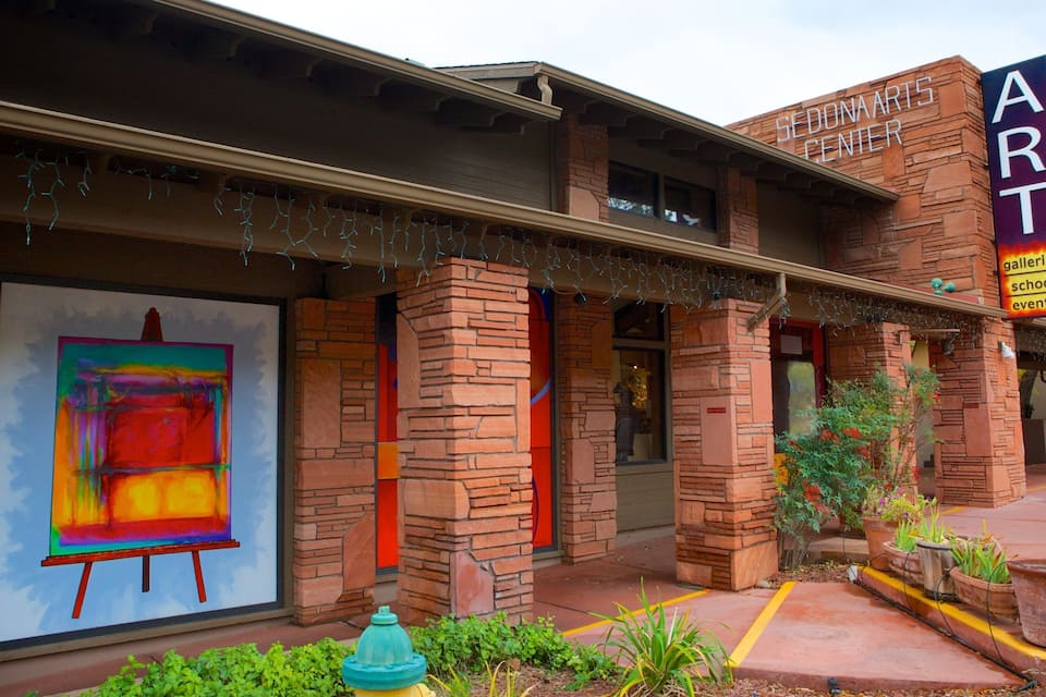 Um edifício com uma pintura abstrata grande e colorida na lateral, fachada de tijolos e uma placa com a inscrição "SEDONA ART GALLERIES SCHOOL EVENTS".