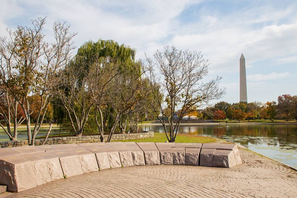Um parque com um lago, árvores, um monumento de pedra e o Monumento a Washington ao fundo.