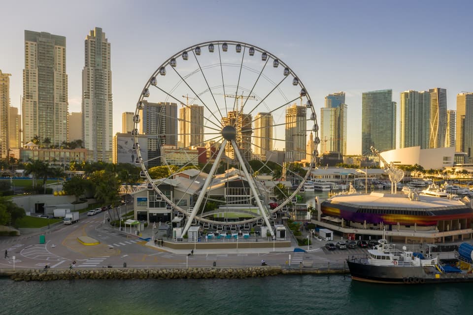 Paisagem urbana à beira-mar com uma grande roda-gigante, edifícios altos modernos e um estádio.