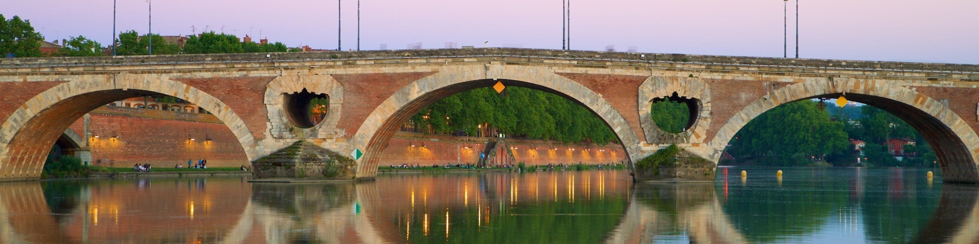 Uma ponte de pedra com três arcos sobre um rio calmo, refletindo a ponte e a vegetação ao redor.