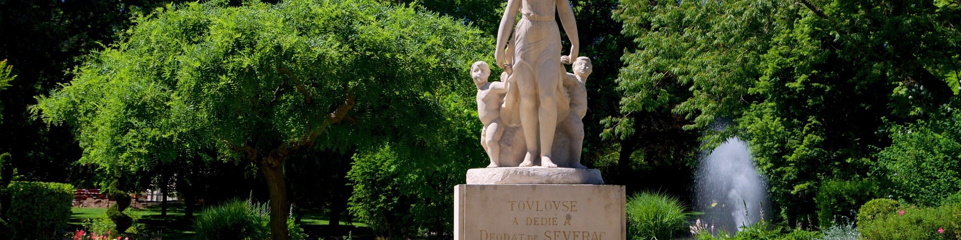 Estátua de uma mulher com uma criança em um parque com vegetação exuberante e uma fonte.