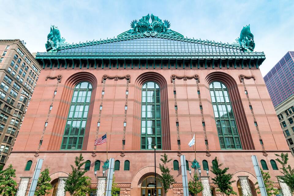 Um edifício grande e histórico com telhado verde, colunas ricamente decoradas e várias bandeiras.