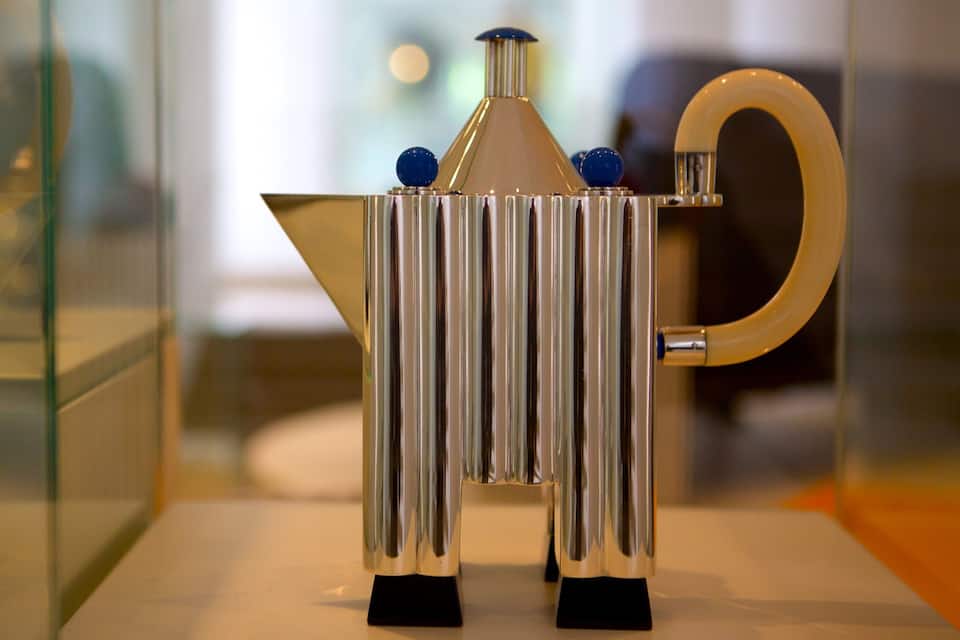 Uma escultura moderna e metálica que se assemelha a uma chaleira, com alça e bico, exposta em uma vitrine de vidro.