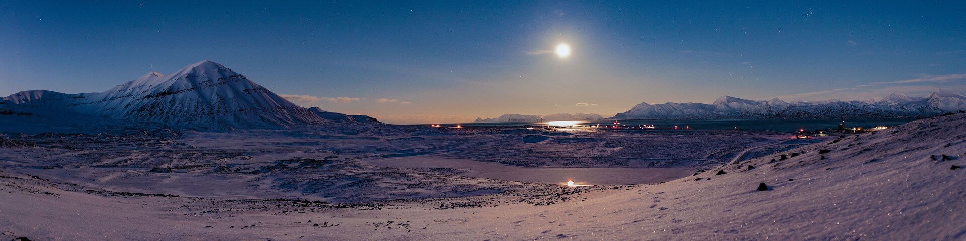 Paisagem nevada de montanhas à noite, com uma lua brilhante e luzes distantes.