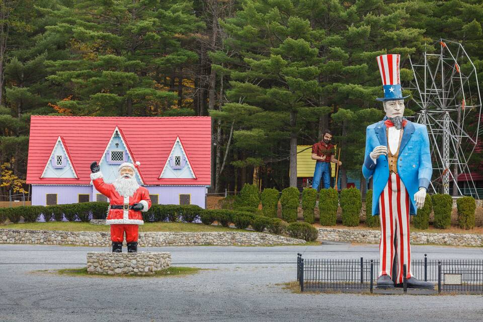 Uma estátua do Papai Noel em frente a uma casa com telhado vermelho e uma roda-gigante ao fundo.