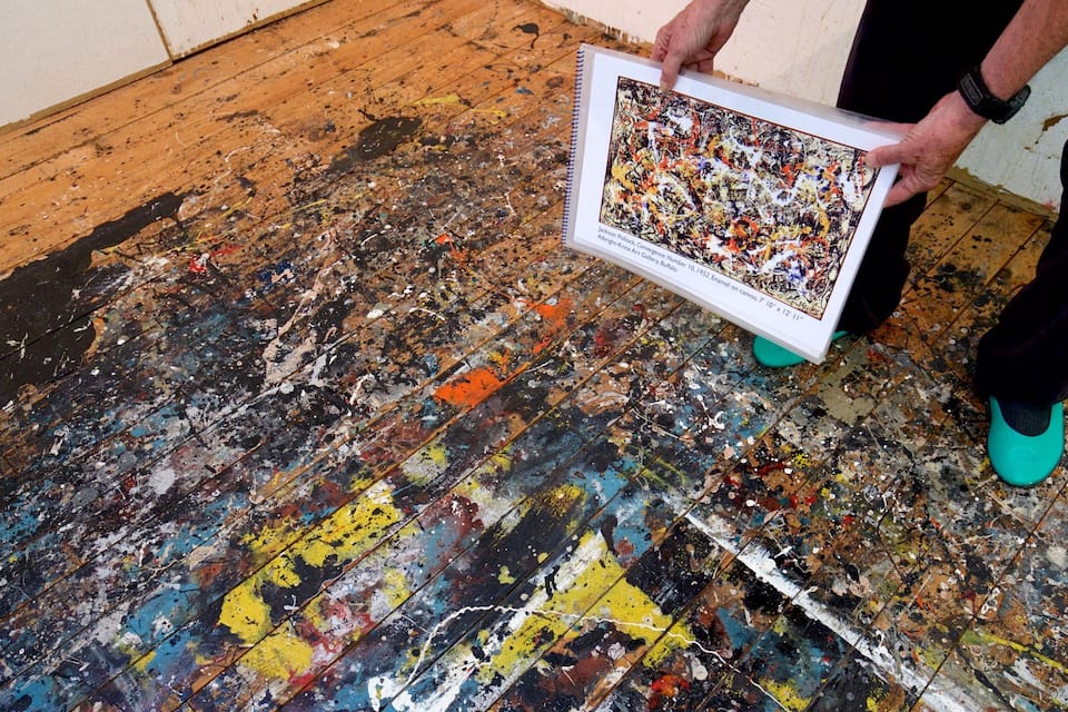Uma pessoa segurando um quadro emoldurado sobre um chão salpicado de tinta.