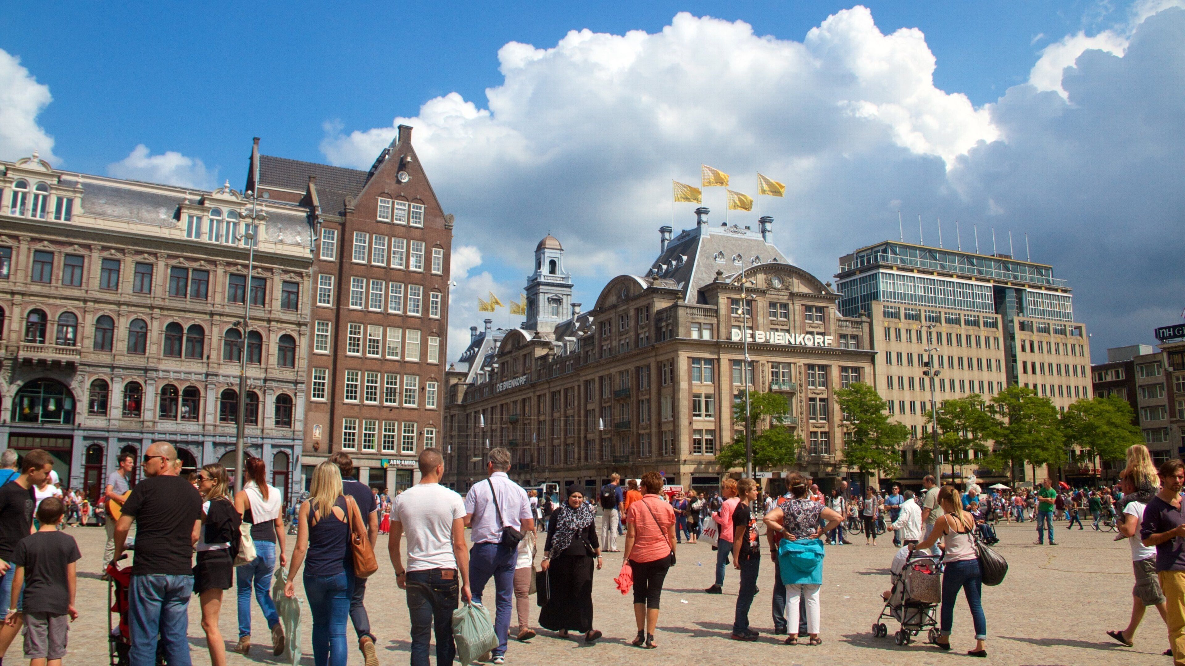 Uma praça pública movimentada com edifícios históricos, incluindo um imponente edifício de tijolos vermelhos com torre do relógio e um edifício moderno de vidro.
