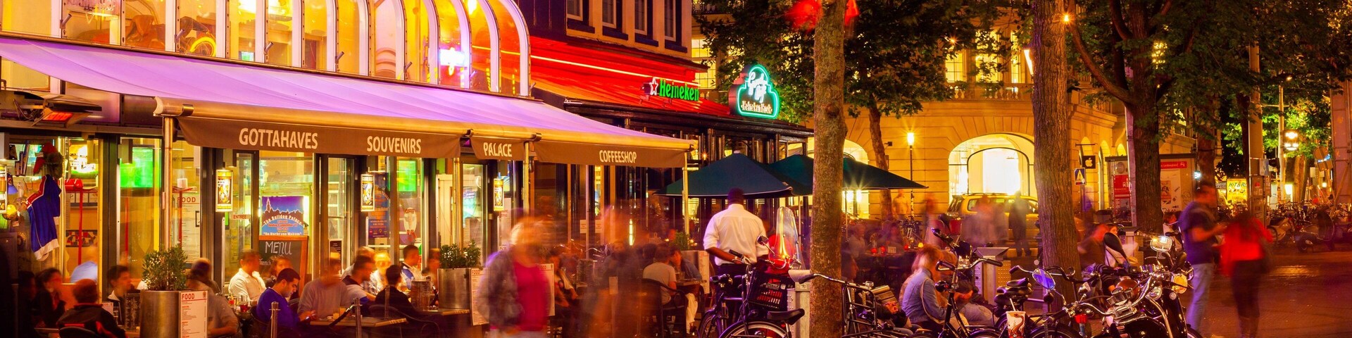 Cena de rua movimentada com diversos estabelecimentos comerciais e cafés, incluindo um anúncio da Heineken e um café chamado 'Palace'.