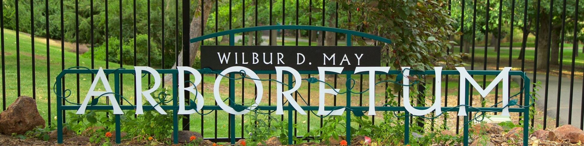 Placa com o nome 'Wilbur D. May Arboretum', cercada por uma grade de metal e plantas floridas.