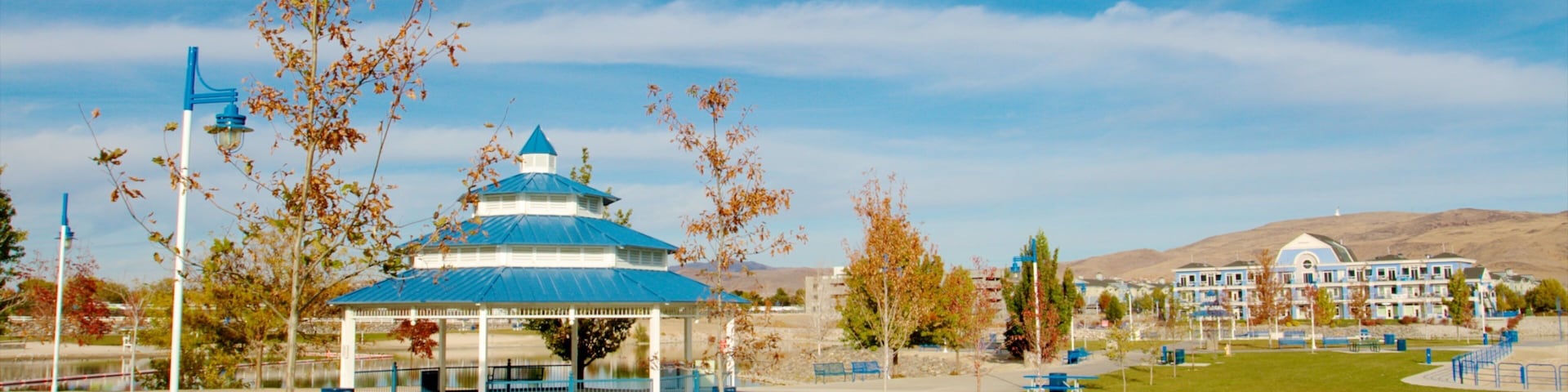Um gazebo com telhado azul, um parque com bancos e um edifício com janelas azuis ao fundo.