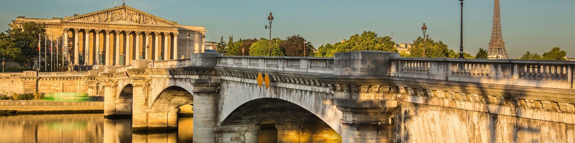 Uma ponte de pedra com arcos sobre um rio calmo, refletindo a arquitetura. Ao fundo, um grande edifício clássico e a Torre Eiffel.