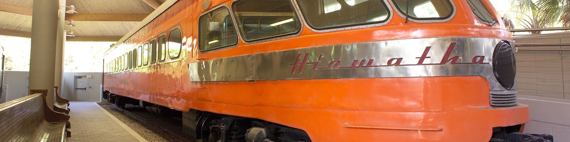 Um trem laranja com a inscrição "Hawatha" na frente, estacionado em uma estação com bancos de madeira e piso de azulejos.