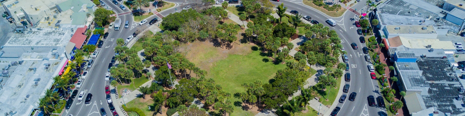 Vista aérea de um cruzamento urbano com uma área verde central, rodeada por estradas e edifícios.