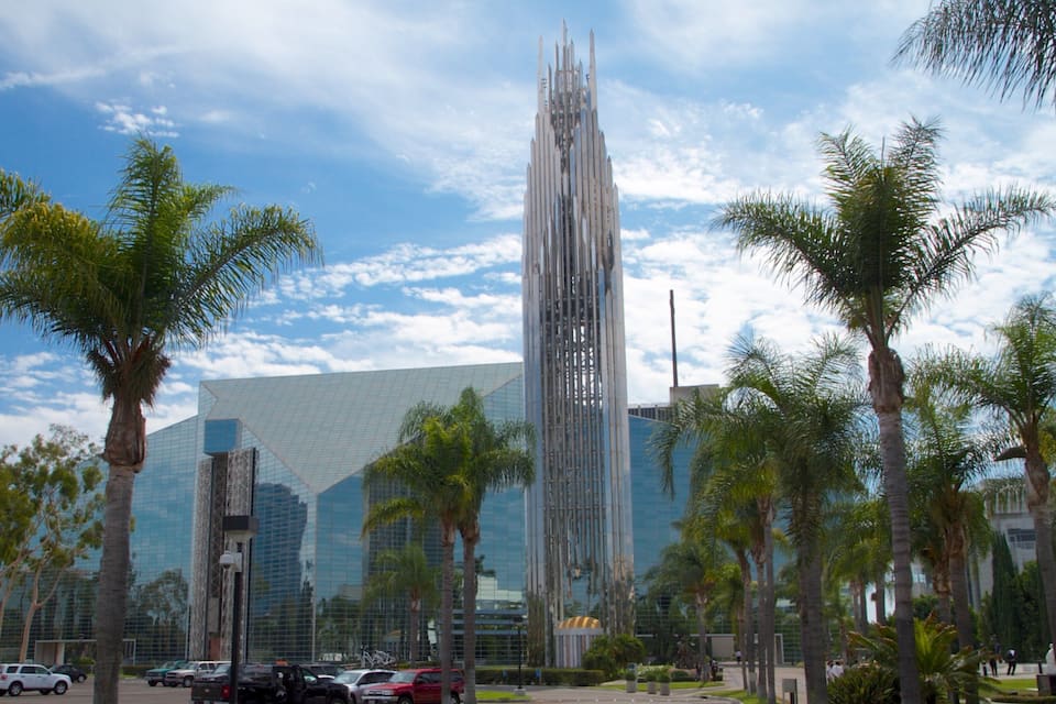 Um arranha-céu moderno com fachada de vidro refletivo, rodeado por palmeiras e outras plantas.