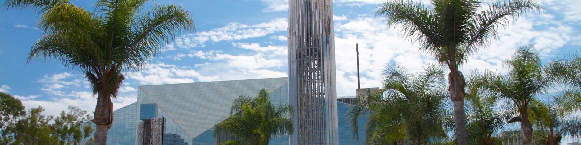 Um arranha-céu moderno com fachada de vidro refletivo, rodeado por palmeiras e outras plantas.