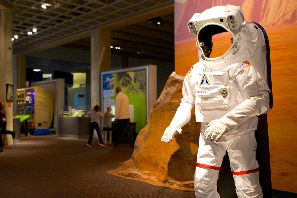 Um manequim de astronauta em tamanho real, vestindo um traje espacial branco com listras vermelhas, em frente a uma grande parede rochosa de cor laranja.