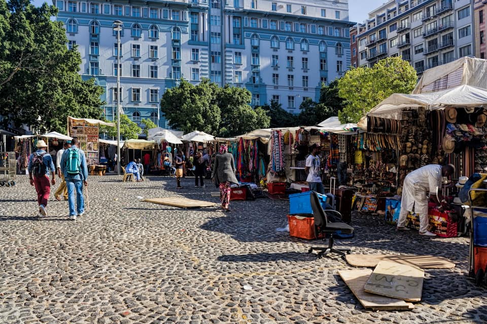 Uma praça de paralelepípedos com bancas de mercado vendendo tecidos e chapéus, cercada por edifícios altos.