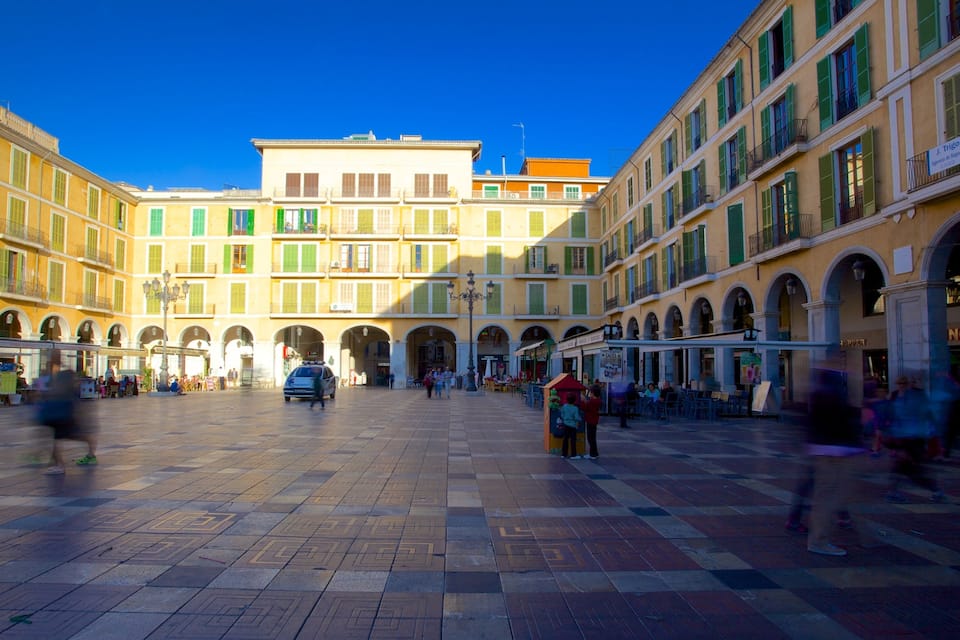 Uma praça com piso de azulejos, edifícios amarelos com persianas verdes e pessoas caminhando.
