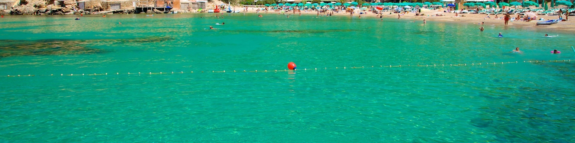 Mar turquesa translúcida com nadadores, uma bóia vermelha e uma praia com guarda-sóis e edifícios.