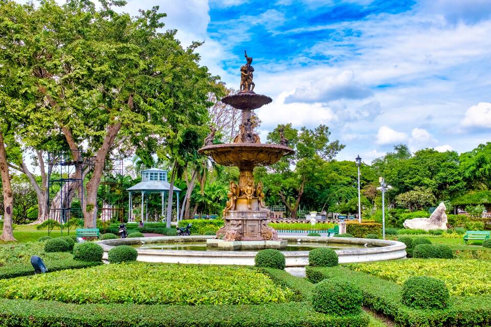 Um parque com uma fonte central, vegetação exuberante e um coreto.