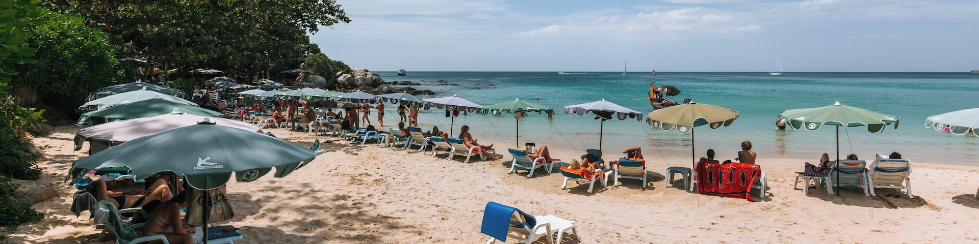 Uma praia com fileiras de espreguiçadeiras e guarda-sóis, pessoas relaxando e barcos na água.