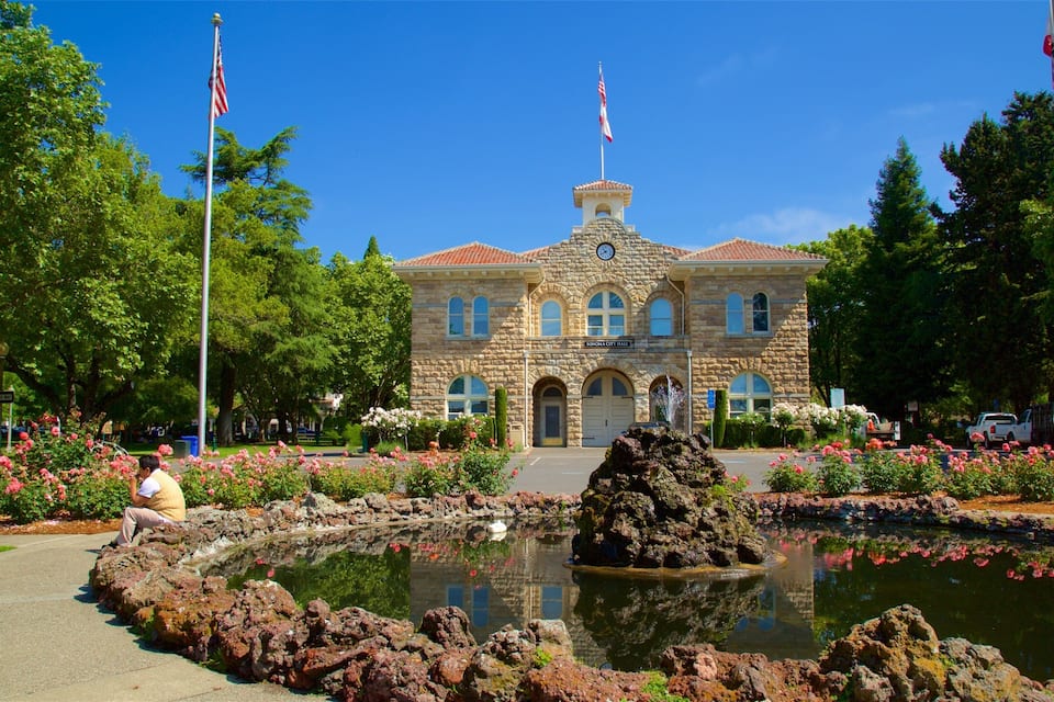 Um edifício de pedra com janelas em arco e uma torre do relógio, rodeado por um jardim com lago e flores.