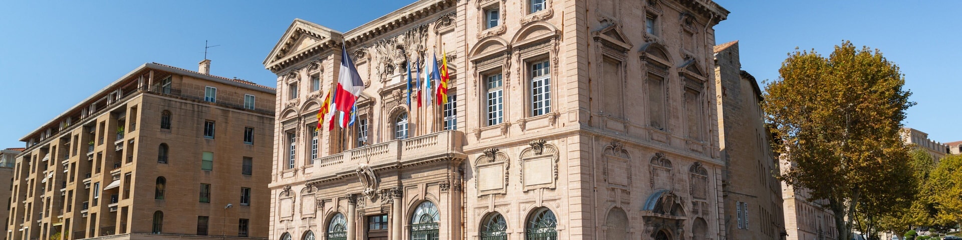 Um edifício histórico com várias bandeiras, amplas janelas e uma praça pavimentada com defensas.