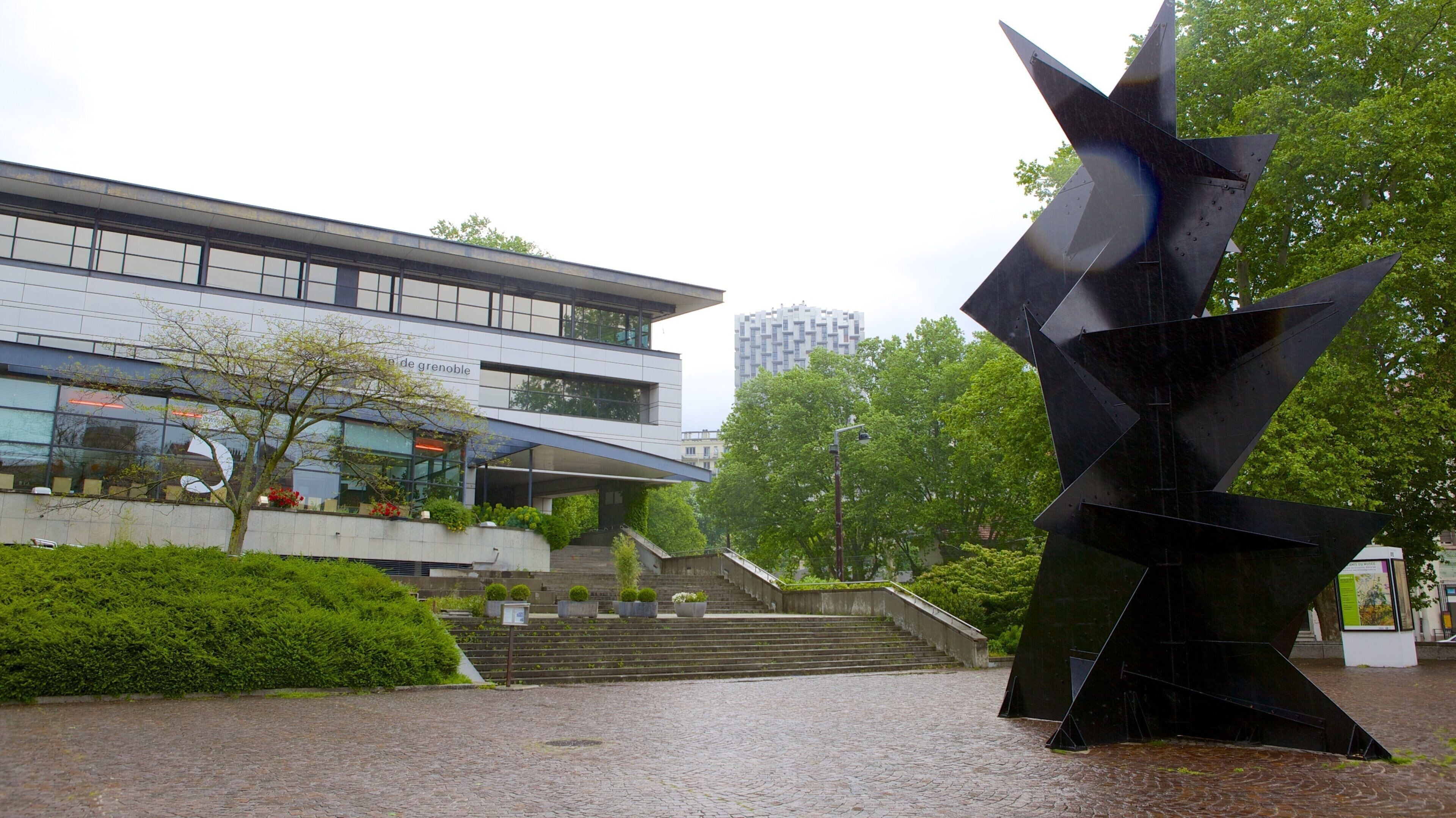 Um edifício moderno com fachada de vidro, uma grande escultura abstrata e uma escadaria que leva à entrada.