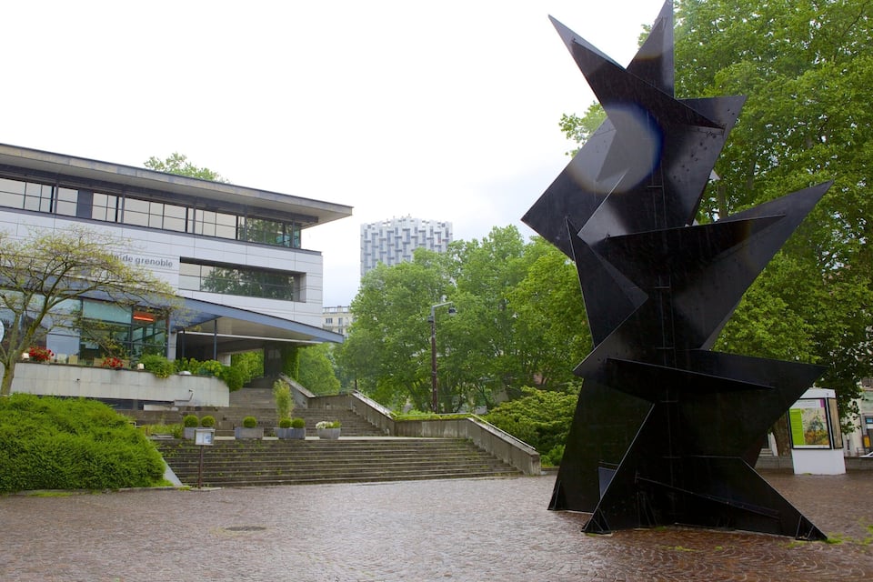 Um edifício moderno com fachada de vidro, uma grande escultura abstrata e uma escadaria que leva à entrada.