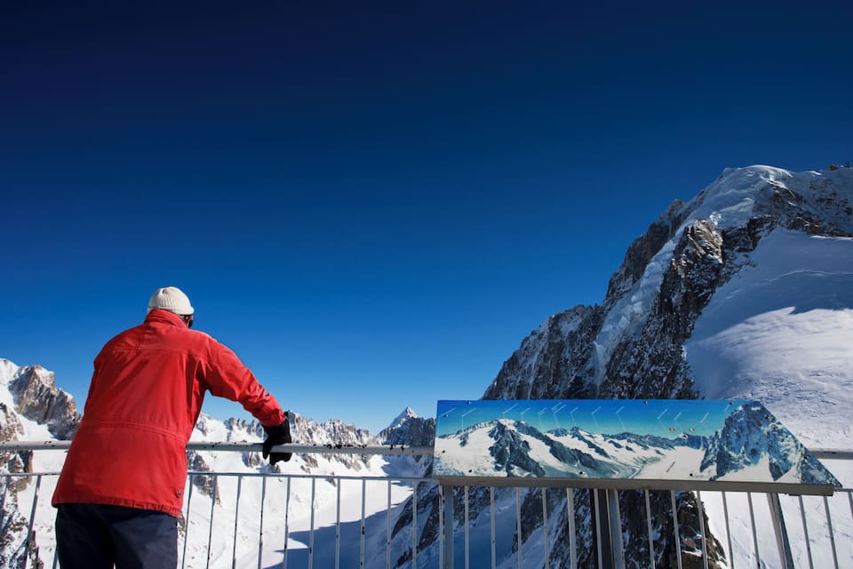 Uma pessoa com casaco vermelho e gorro branco, em frente a um corrimão, com vista para uma cadeia montanhosa coberta de neve e um céu azul límpido.