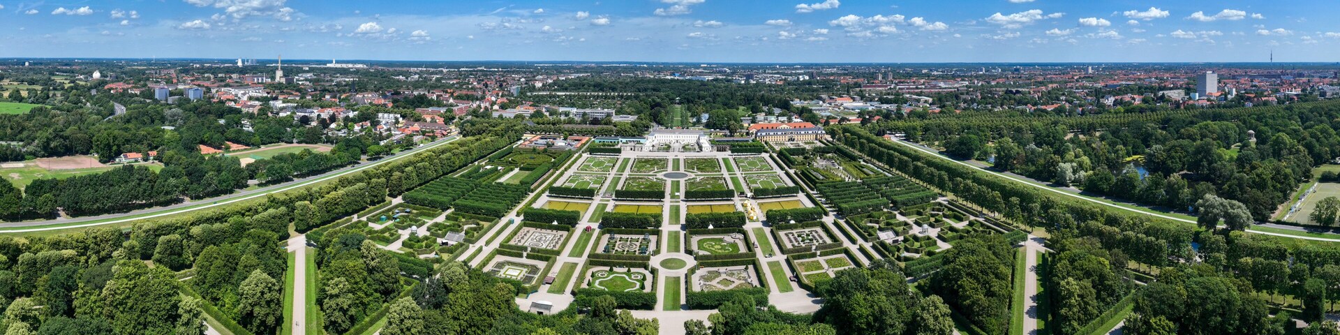 Um jardim amplo e simétrico, com gramados bem cuidados e fontes, cercado por vegetação densa e edifícios urbanos ao fundo.