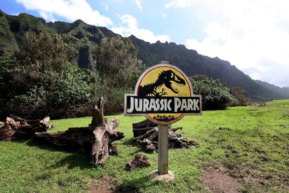 Placa para Jurassic Park com silhueta de dinossauro, ao fundo colinas verdes exuberantes e céu claro.