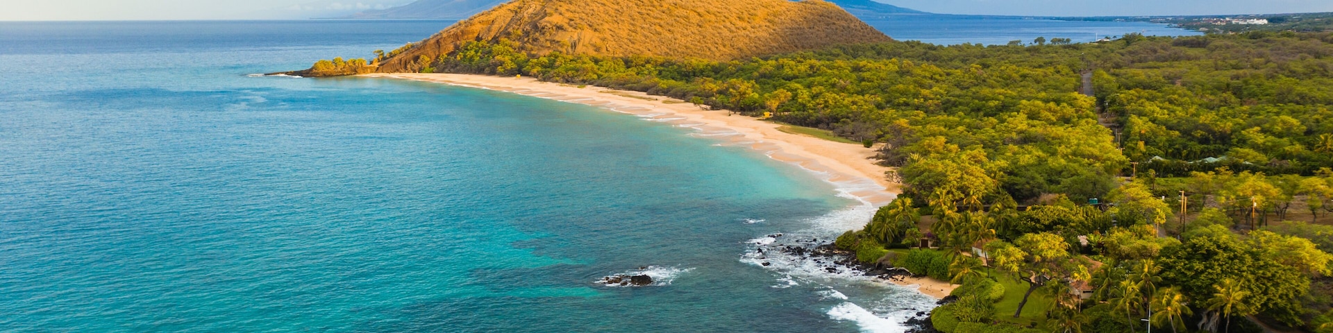 Paisagem costeira com praia arenosa, águas verde-turquesa e uma colina coberta por vegetação densa.