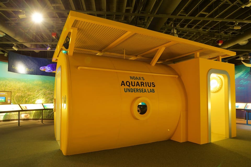 Uma exposição amarela identificada como 'Laboratório Submarino AQUARIUS da NOAA', com uma janela circular e uma porta.