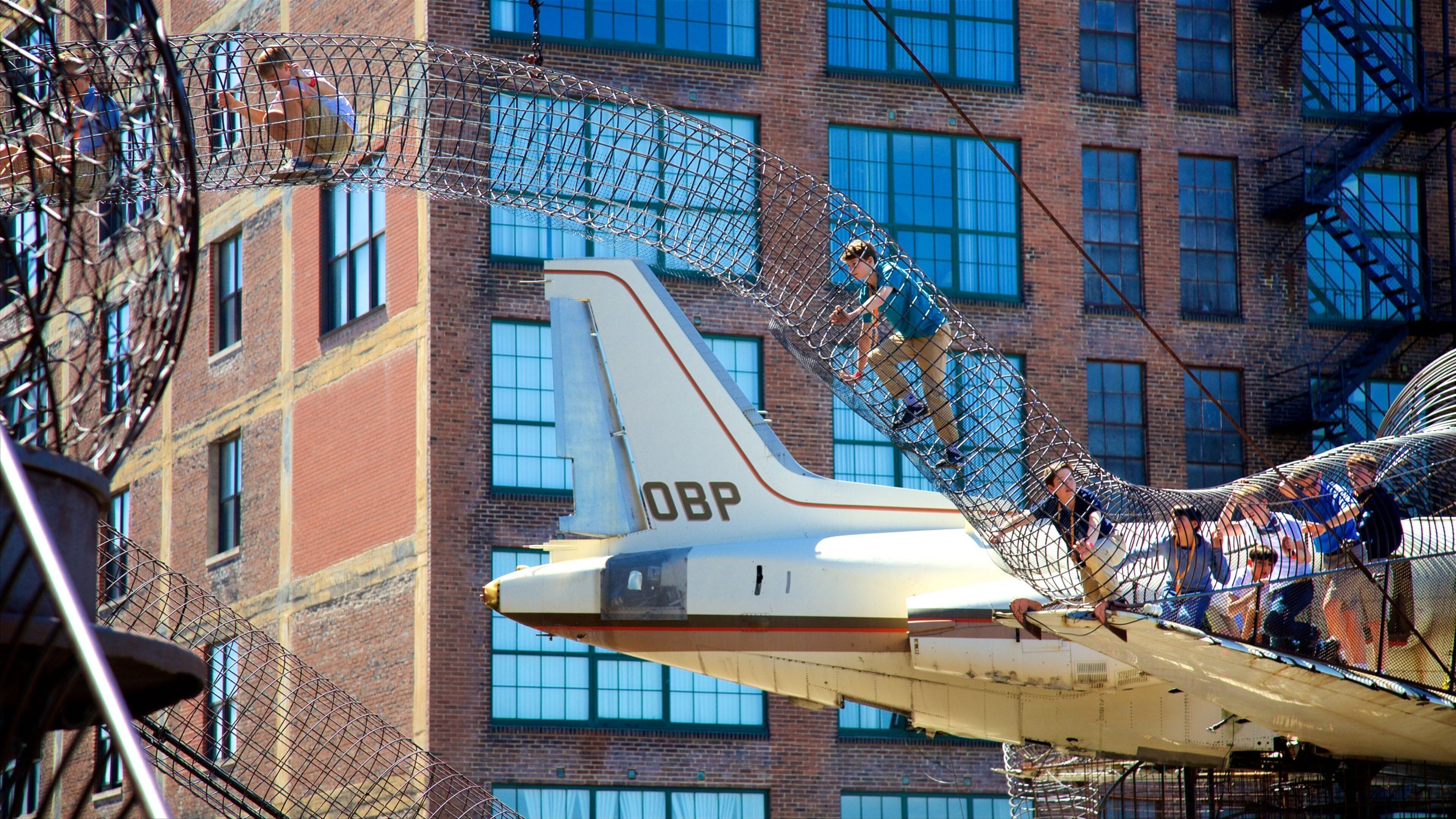 Um avião antigo com a matrícula OBP, suspenso em uma estrutura de rede, com um edifício de tijolos e várias janelas ao fundo.