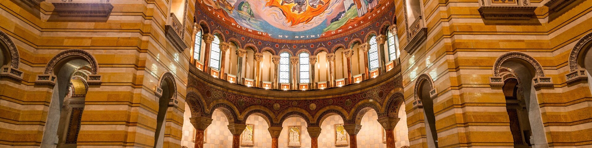 Interior de igreja com um grande afresco ornamentado no teto, janelas em arco e fileiras de bancos de madeira.