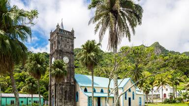 Uma igreja histórica com uma torre do relógio, uma pequena capela e vegetação exuberante.
