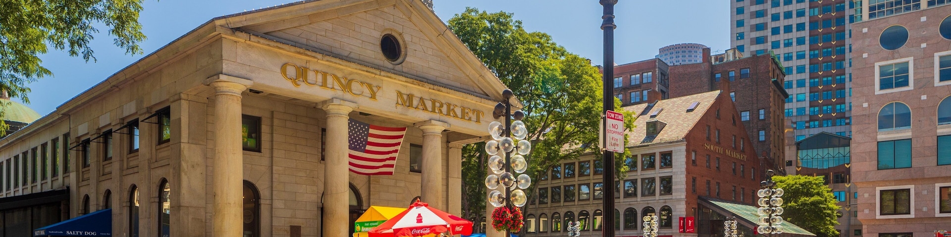 Mercado Quincy, com uma torre do relógio em destaque, bandeira americana e área de vendedores ambulantes.