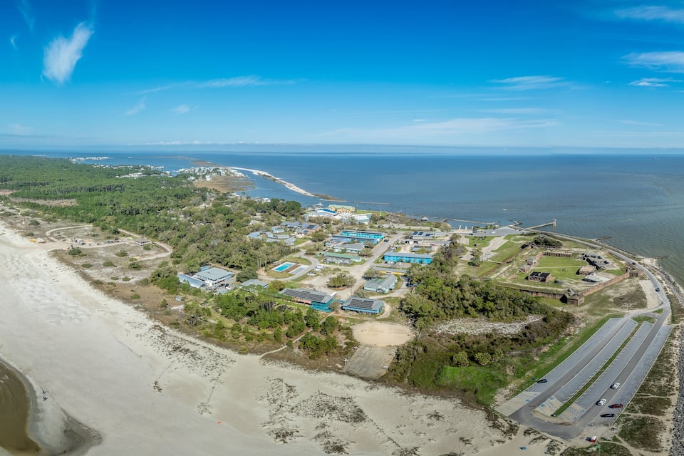 Vista aérea de uma área costeira com uma praia, uma estrada e um conjunto de edifícios.