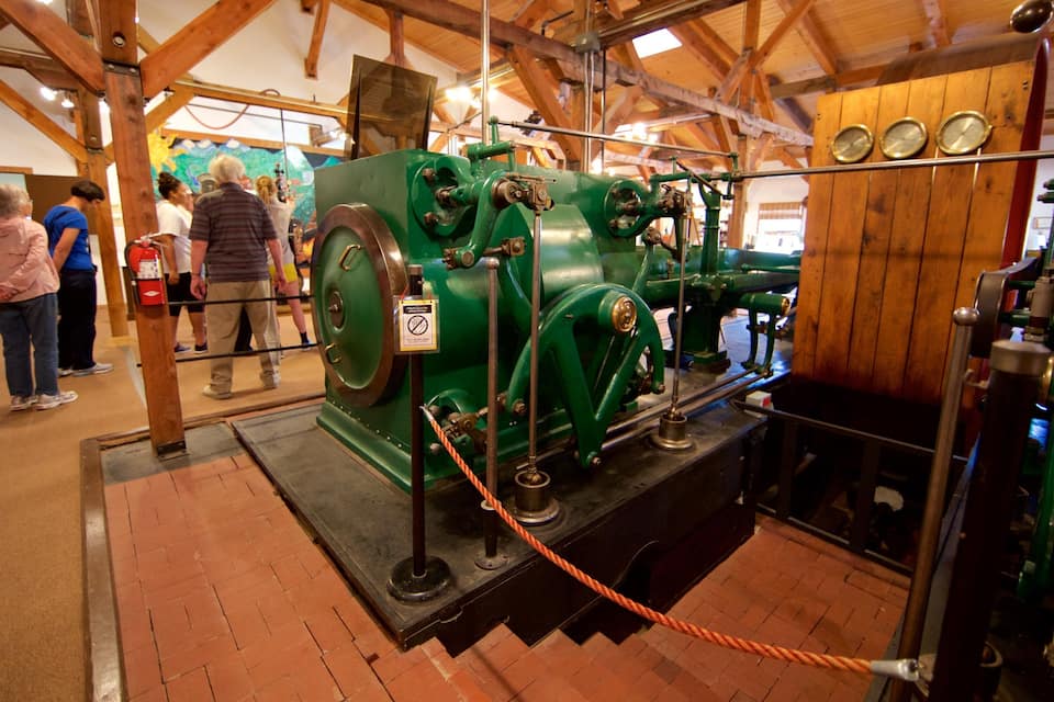 Uma máquina industrial verde grande com manômetros e tubulações, rodeada por um chão de tijolos e vigas de madeira.