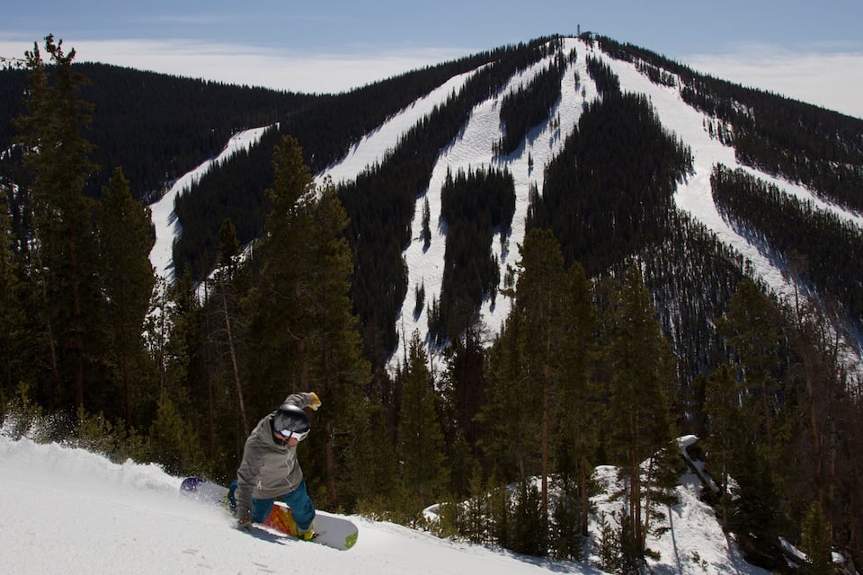 Um snowboarder numa encosta nevada de montanha, com uma paisagem arborizada ao fundo e céu claro.