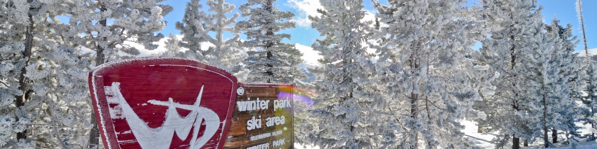 Placa de área de esqui coberta por neve, com um símbolo de esqui, rodeada por árvores carregadas de neve e um céu azul claro.