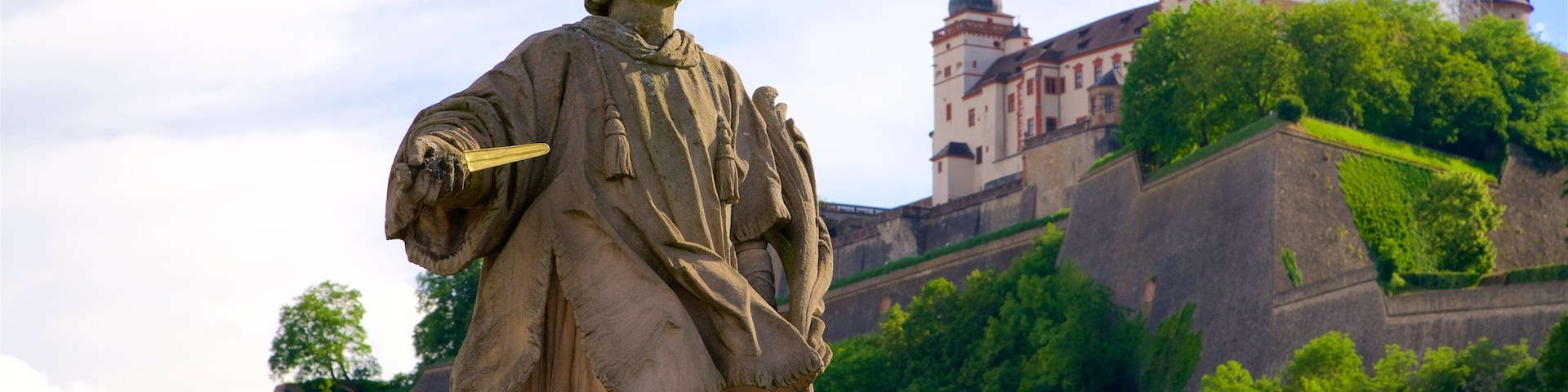 Uma estátua de uma figura histórica segurando uma espada, com um castelo e vegetação exuberante ao fundo.